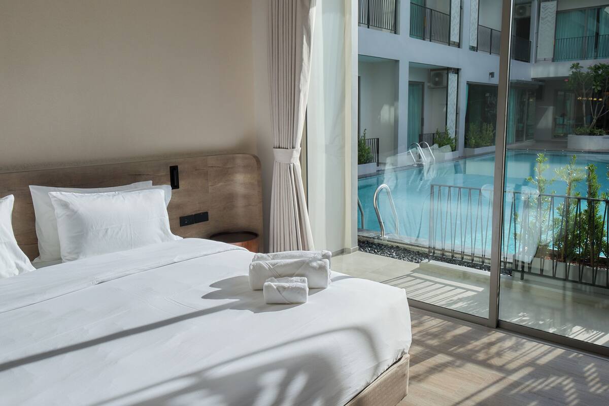 Quarto de hotel com uma cama grande, roupa de cama branca, vista para uma piscina e uma varanda com plantas em vasos.
