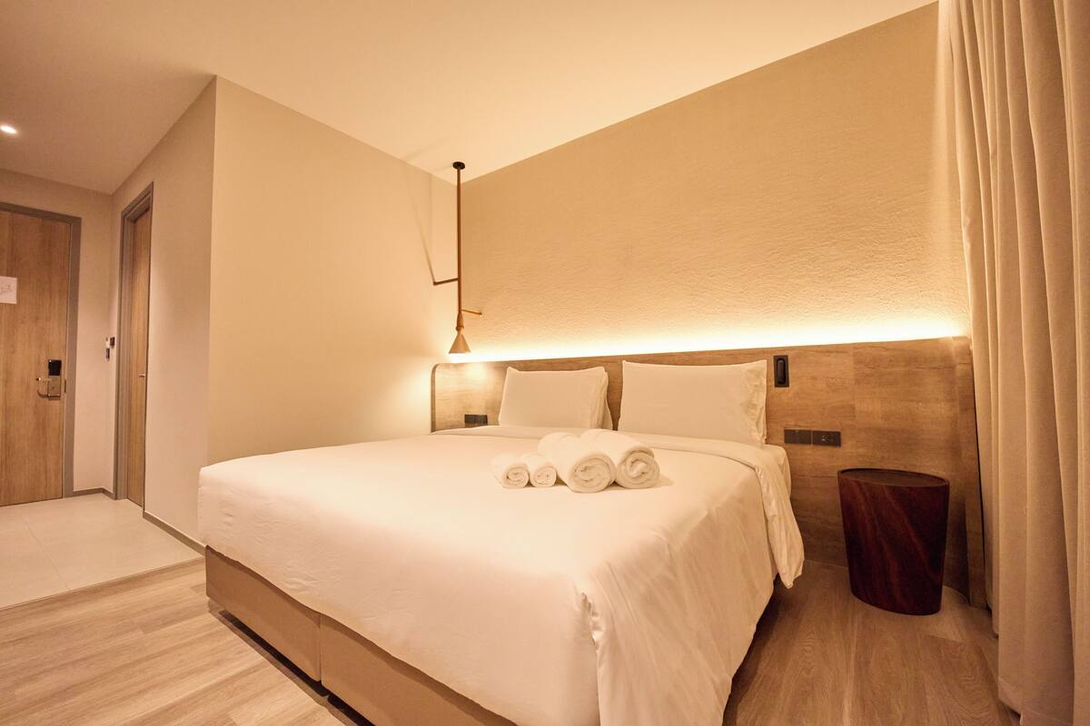 Quarto de hotel moderno com uma cama grande, mesa de cabeceira, abajur e piso de madeira.