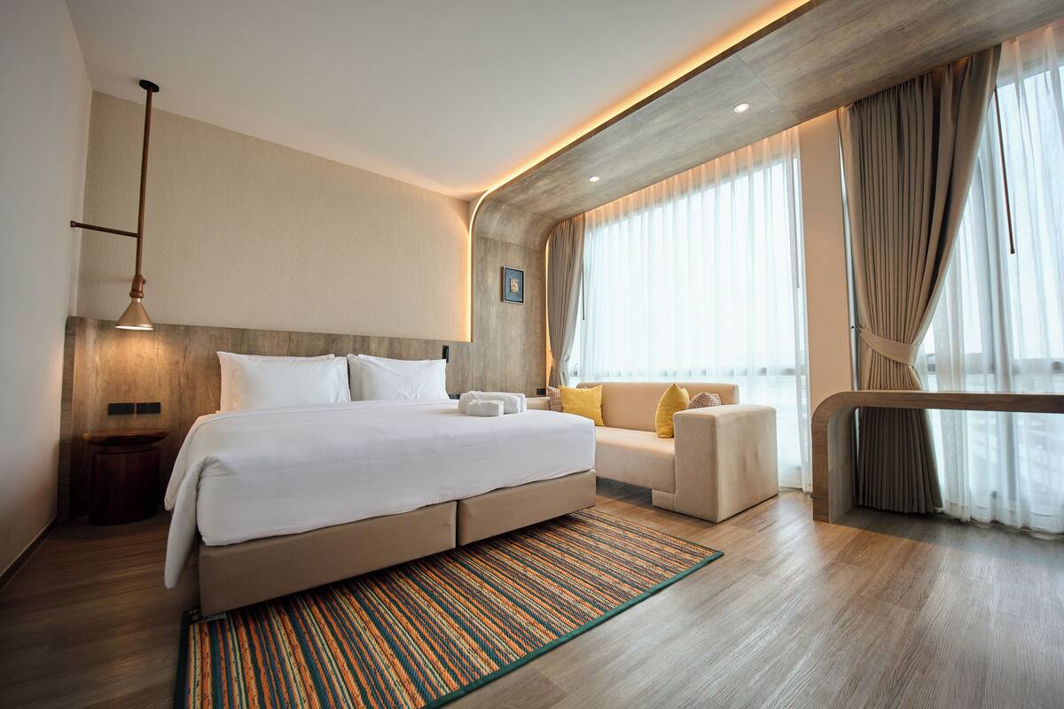 Quarto de hotel moderno com uma cama grande, um sofá, uma pequena mesa e vista da cidade através de amplas janelas.
