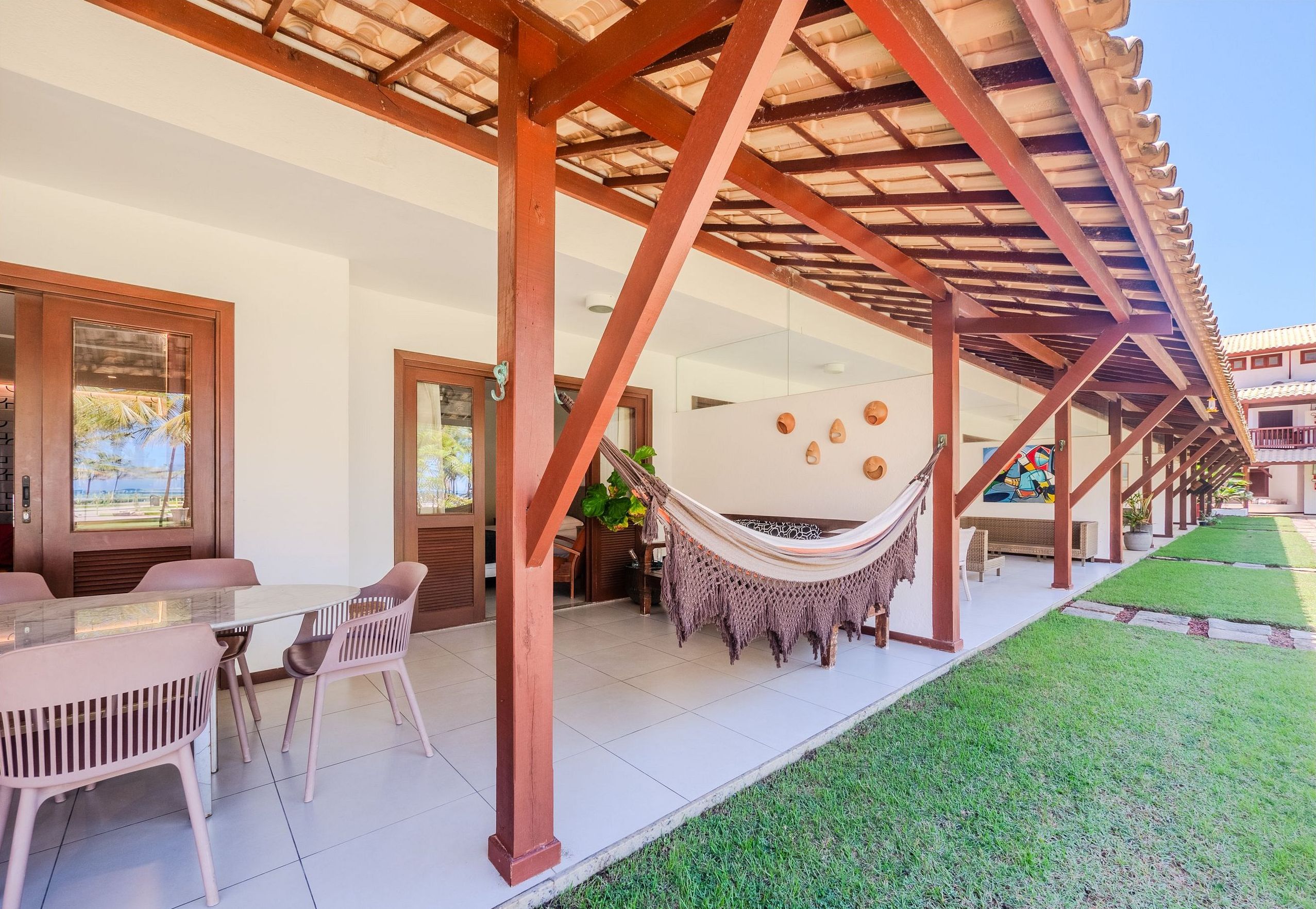 Uma área de estar coberta ao ar livre, com uma rede, uma mesa de jantar com cadeiras e vista para o mar através de uma janela.