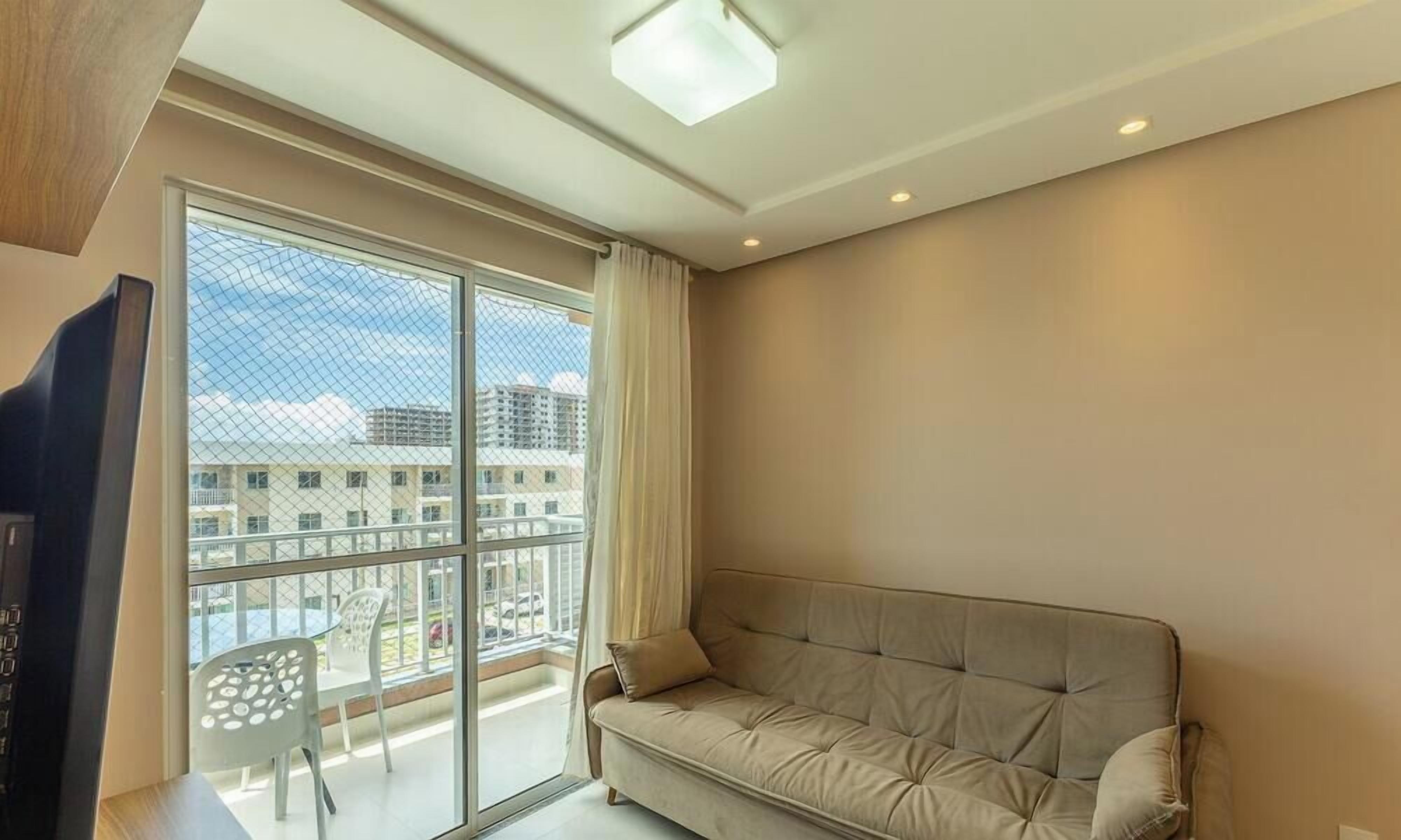 Uma sala de estar moderna com um sofá, uma mesinha e uma cadeira. Há uma janela grande com vista para edifícios.