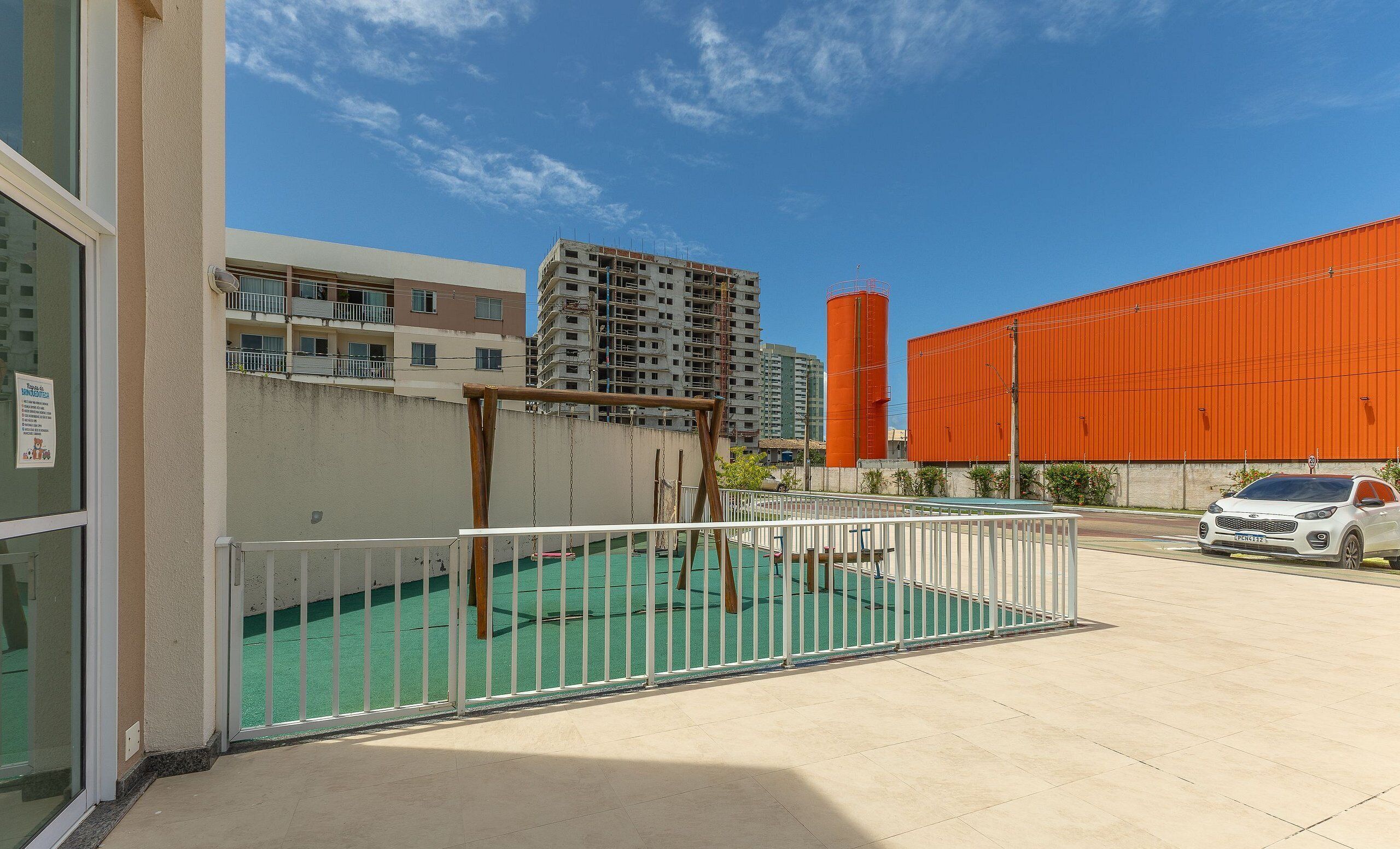 Um edifício residencial com área externa cercada, piscina e um grande edifício industrial laranja ao fundo.