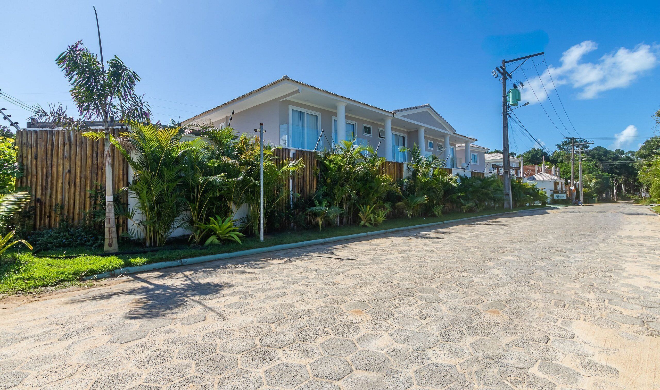 Rua residencial com uma casa, telhado em tijolos, cerca de madeira e vegetação exuberante.