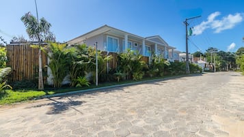 Rua residencial com uma casa, telhado em tijolos, cerca de madeira e vegetação exuberante.