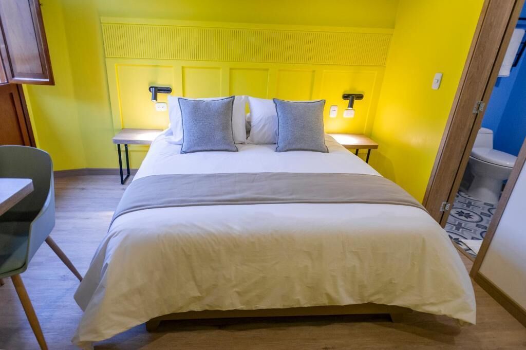 Quarto de hotel com cama, mesa de cabeceira, cadeira e sanita.