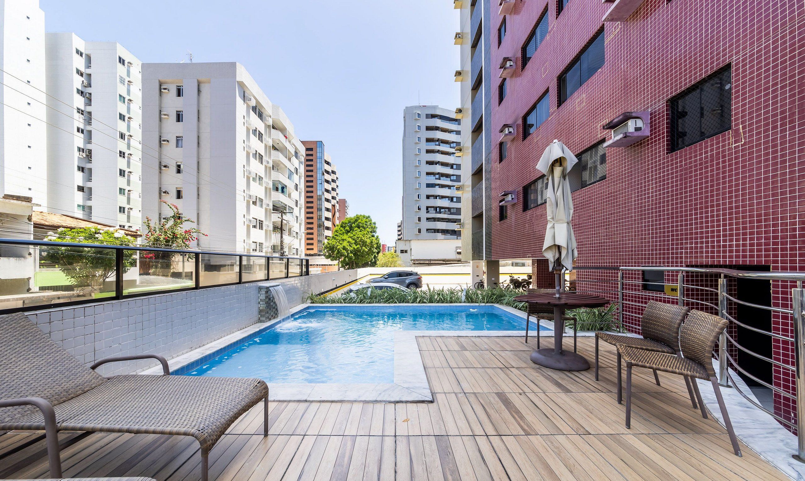 Uma área de piscina no terraço com um deck de madeira, espreguiçadeiras e um edifício com telhado vermelho ao fundo.