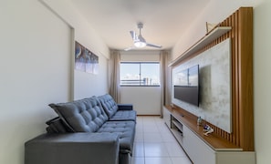 Uma sala de estar moderna com um sofá cinzento, um ventilador de teto, uma TV fixada num painel de madeira e uma janela com vista para edifícios.