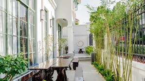 Um pátio com área de refeições, plantas de bambu e um edifício com paredes brancas e molduras de janela verdes.