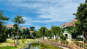 Um lago tranquilo com uma ponte de madeira, cercado por vegetação exuberante e edifícios tradicionais com telhados vermelhos.