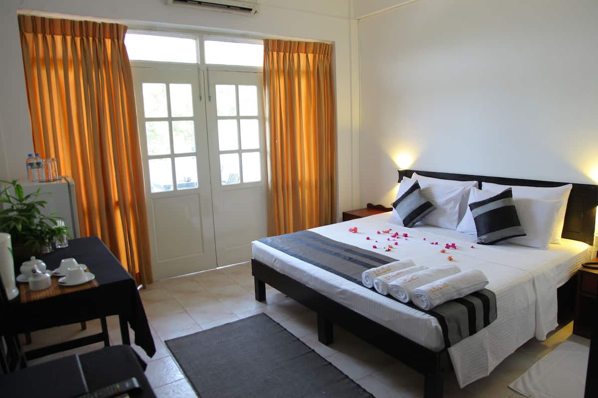 Quarto de hotel bem arrumado, com cama, mesas de cabeceira, escrivaninha com cadeira, planta em vaso e janela com cortinas.
