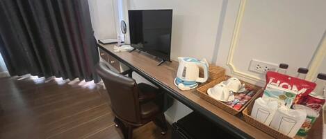 Uma pequena cozinha compacta com geladeira, cafeteira e micro-ondas. Há uma cadeira e uma escrivaninha com um computador.