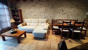 Uma sala de estar acolhedora com uma parede de pedra, um sof á branco, uma mesa de jantar de madeira com cadeiras e um chão ladrilhado.