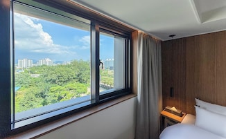 Quarto de hotel com uma grande janela que oferece vista para a cidade, uma cama e uma mesa de cabeceira com luminária.