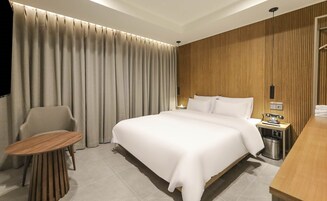 Quarto de hotel com uma cama grande, uma cadeira, uma mesinha e um criado-mudo com abajur e telefone.