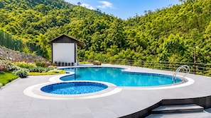 Piscina com um pequeno edifício, cercada por vegetação exuberante e montanhas ao fundo.