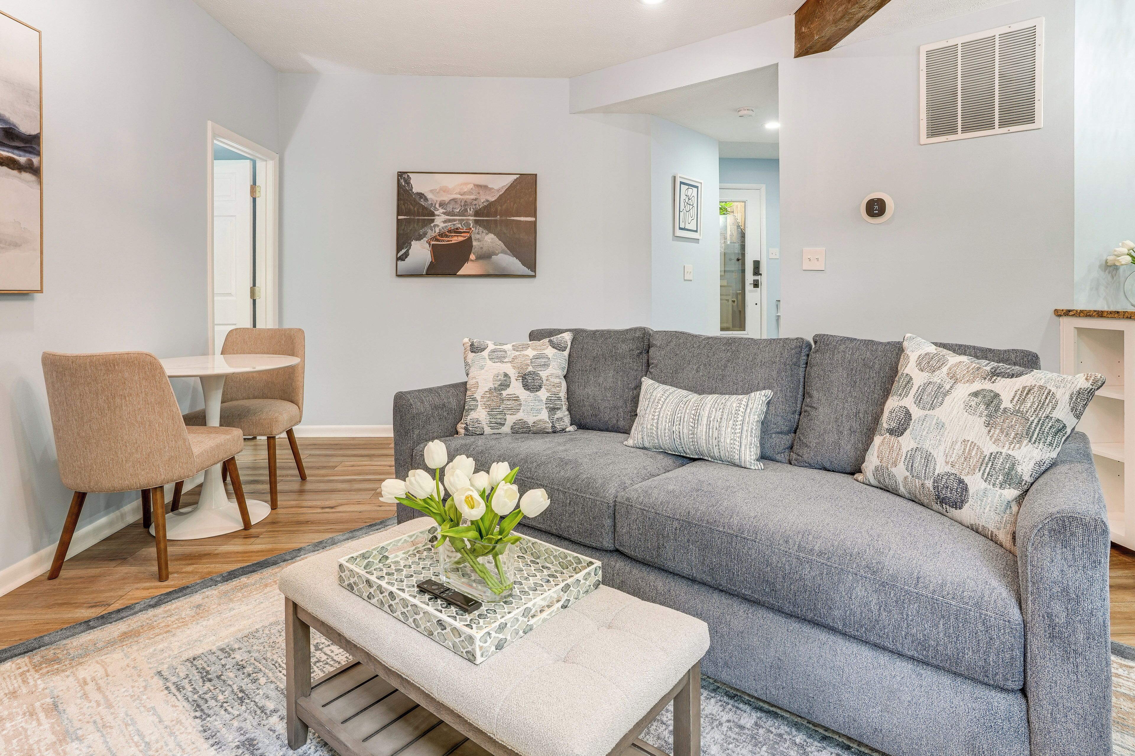 Uma sala de estar moderna com um sofá cinzento, almofadas estampadas, uma mesa de centro com um vaso de flores, e um espaço de jantar com cadeiras e uma mesa.