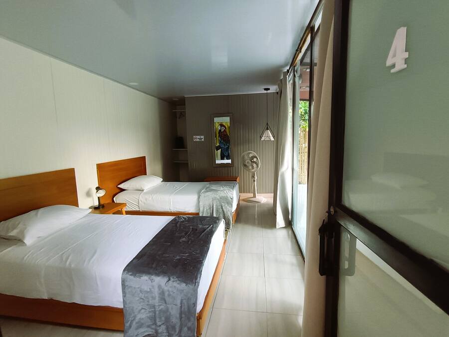 Quarto de hotel com duas camas, uma mesa de cabeceira com abajur, um ventilador de teto e uma janela com cortinas.