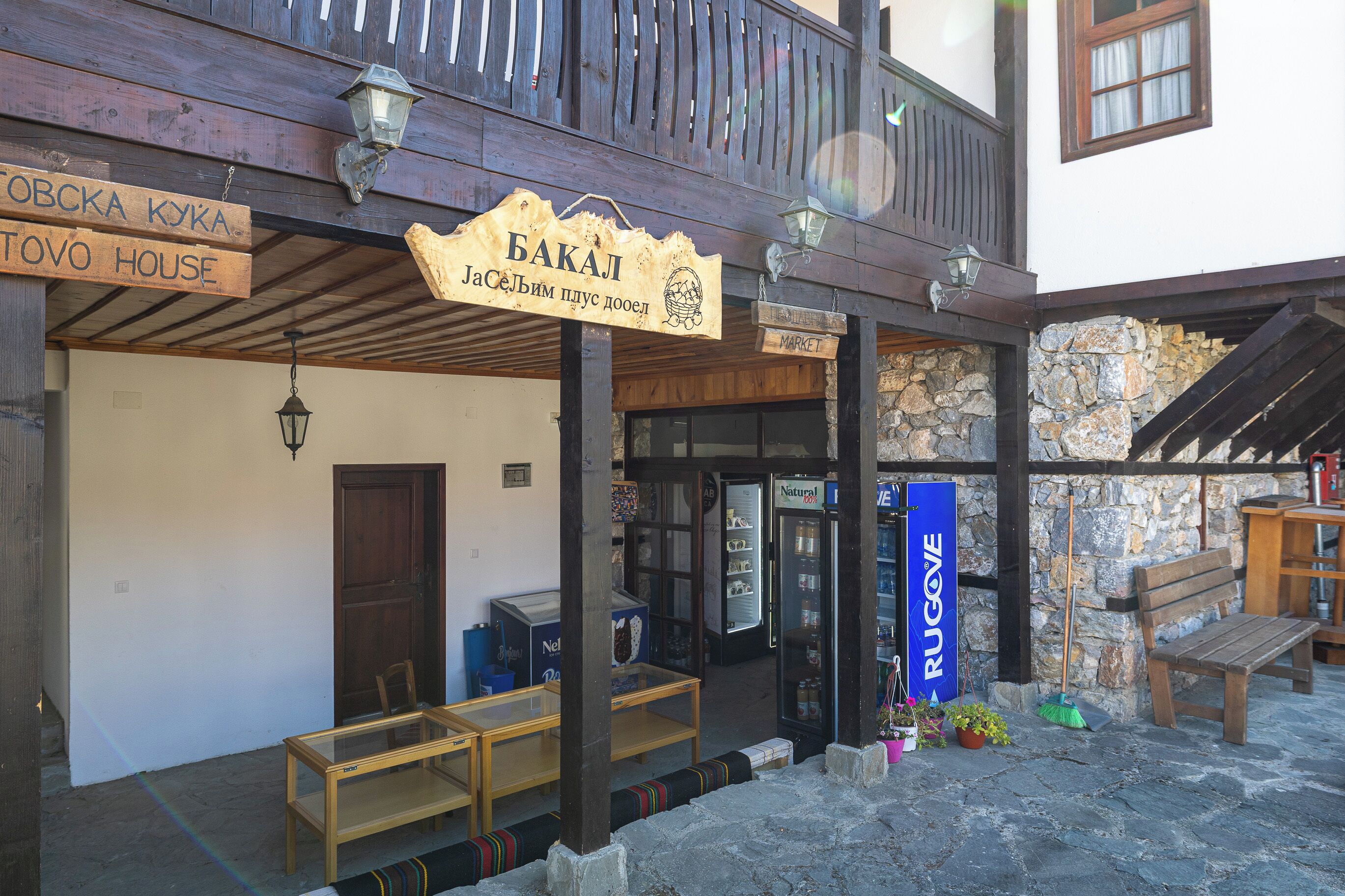 Um edifício de madeira com uma placa que diz 'Bakal' e 'Mercado'. Há bancos do lado de fora e um muro de pedra ao fundo.