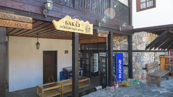 Um edifício de madeira com uma placa que diz 'Bakal' e 'Mercado'. Há bancos do lado de fora e um muro de pedra ao fundo.