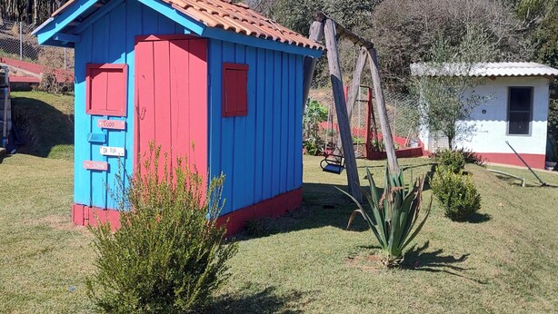 Uma pequena estrutura de madeira colorida, com uma porta vermelha e telhado azul, cercada por grama e árvores.