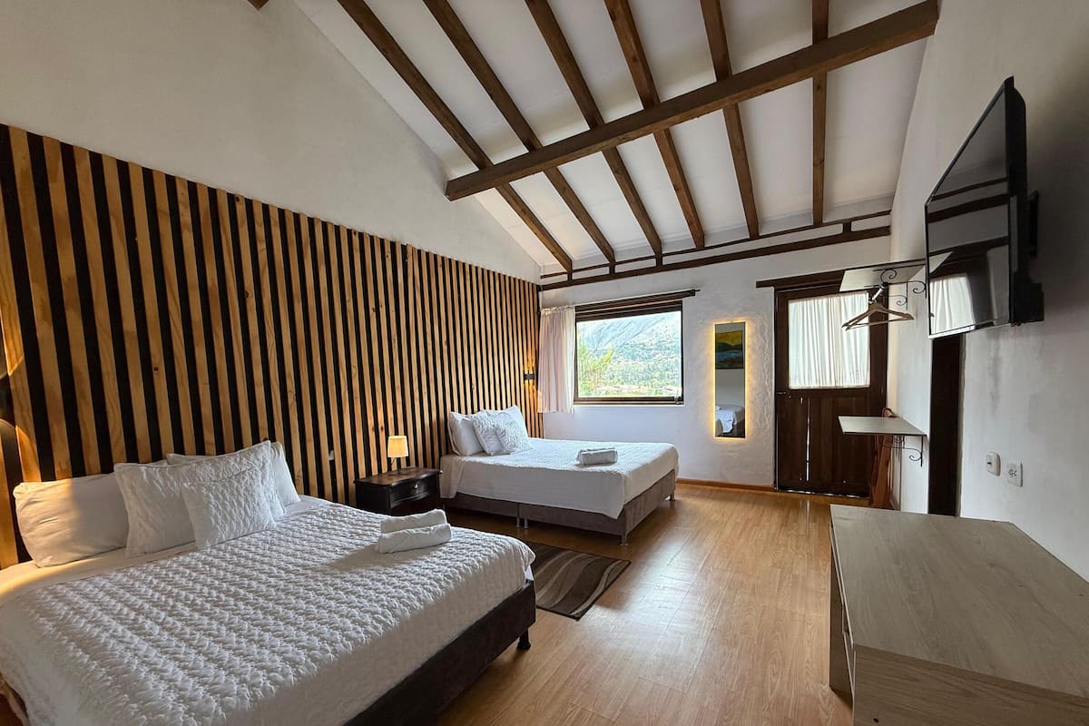 Quarto com duas camas, paredes de ripas de madeira, teto com vigas aparentes, janela com vista, mesa de cabeceira com abajur e TV na parede.