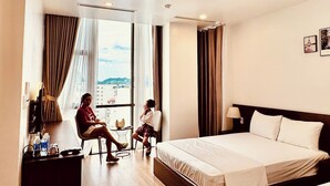Quarto de hotel com uma cama grande, uma mesa e uma cadeira. Há duas pessoas no quarto, uma sentada e outra em pé.