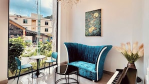 Um quarto espaçoso com um piano, um sofá azul, um candelabro e uma grande janela que oferece vista para edifícios e áreas verdes.
