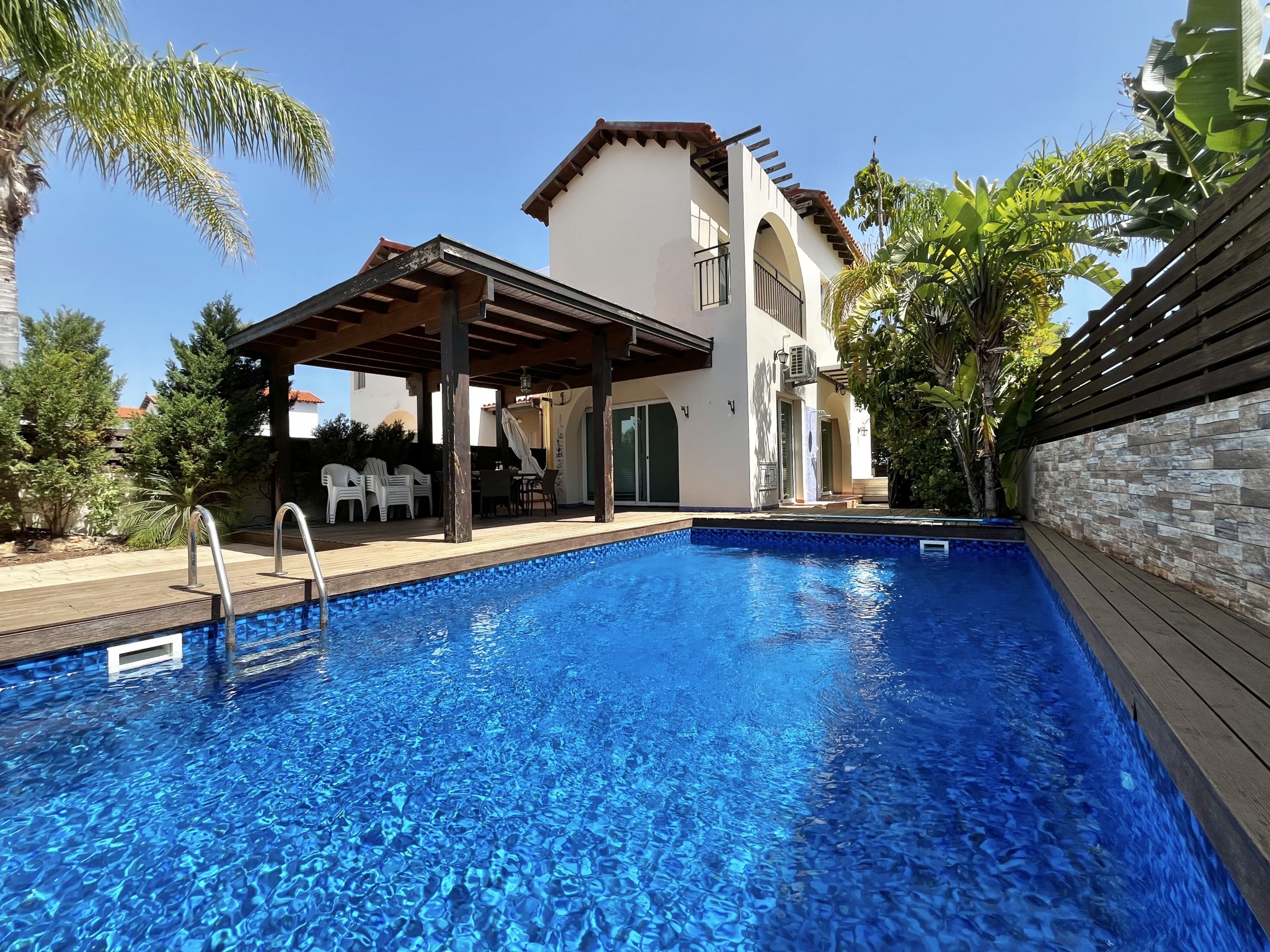 Piscina com deck de madeira, área de varanda coberta e uma casa de dois andares com fachada de pedra.