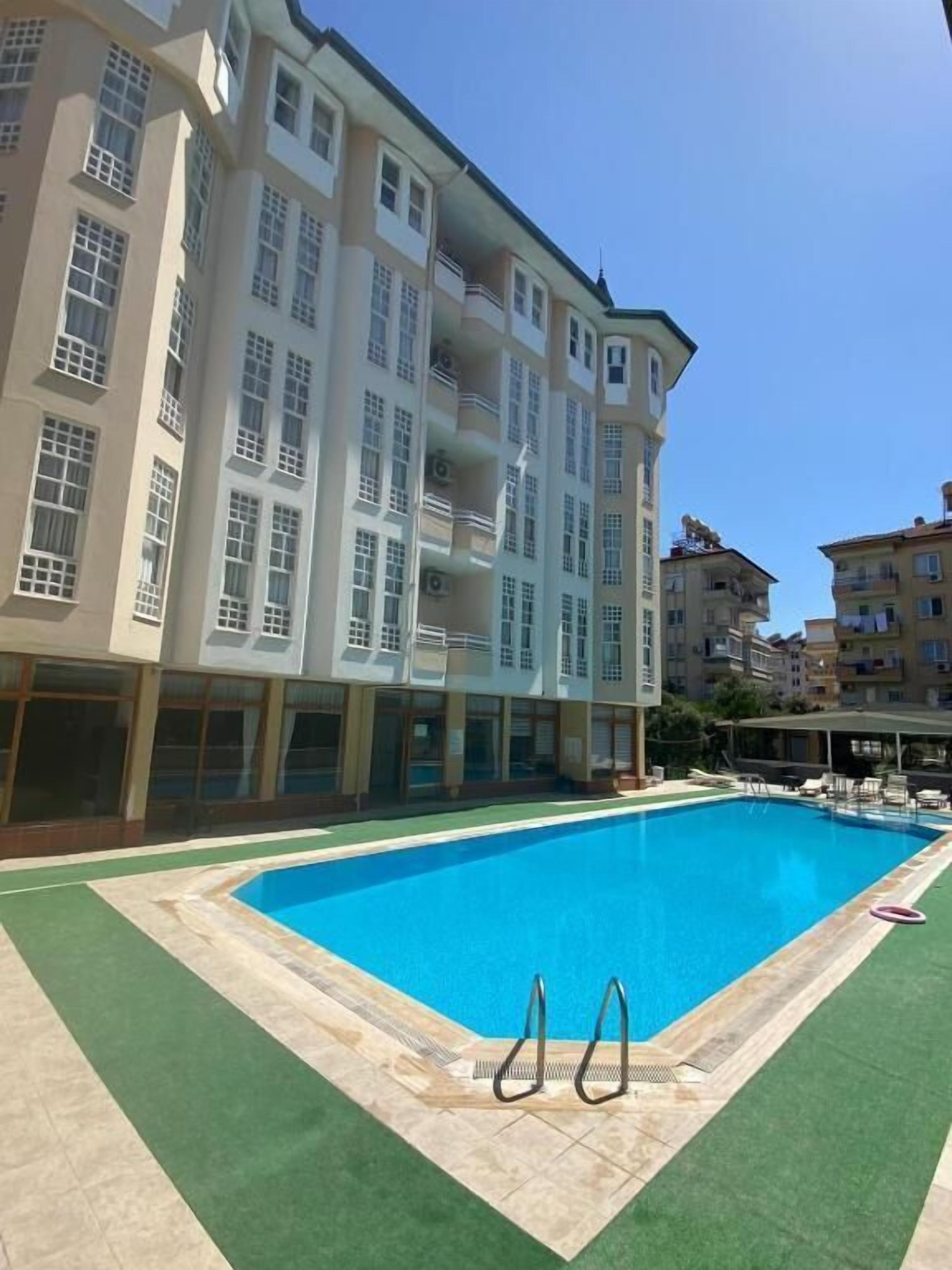 Piscina com borda verde e duas plataformas de mergulho, cercada por um edifício com várias varandas e janelas.