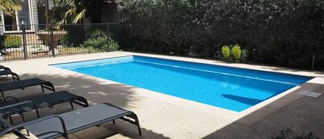 Uma piscina retangular com superfície de água azul cristalina, rodeada por um deck de concreto. Várias espreguiçadeiras estão dispostas ao longo da borda da piscina.