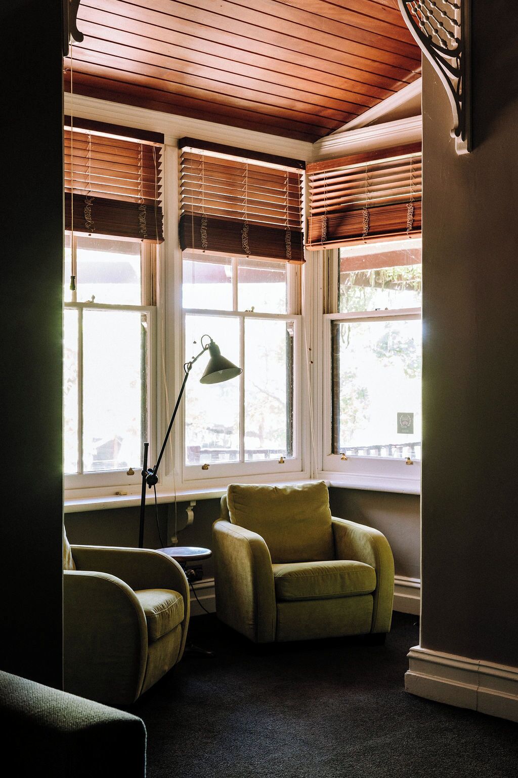 Quarto nos cantos com janela saliente, uma poltrona verde, luminária de chão e uma pequena mesa.