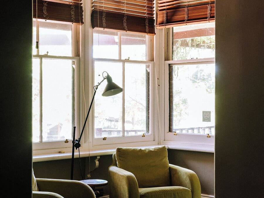 Quarto nos cantos com janela saliente, uma poltrona verde, luminária de chão e uma pequena mesa.
