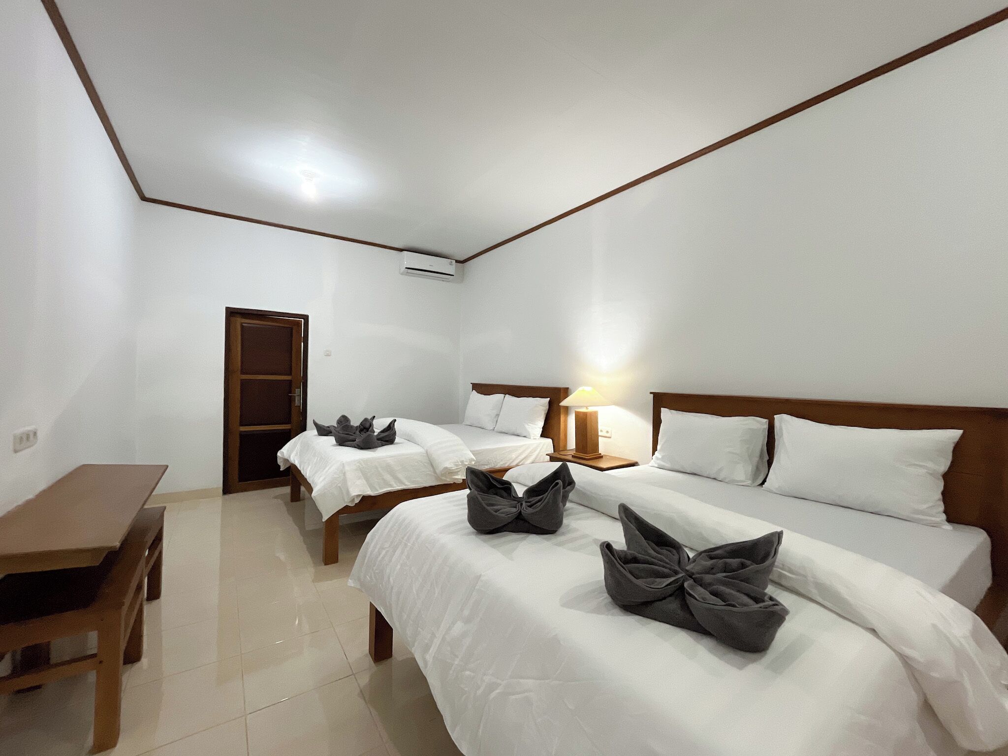 Quarto de hotel com duas camas, uma mesa de madeira e um criado-mudo com abajur.