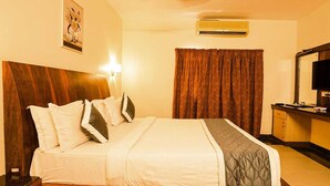Quarto de hotel com cama, escrivaninha, televisão, ar condicionado e ventoinha de teto.