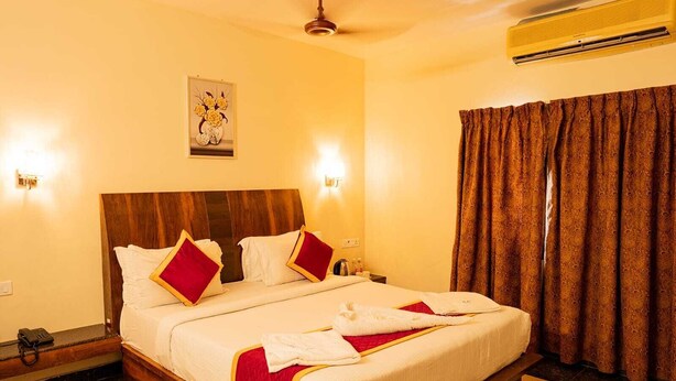 Quarto de hotel com uma cama grande, duas mesas de cabeceira, um ventilador de teto, um aparelho de ar condicionado e uma janela com cortinas.