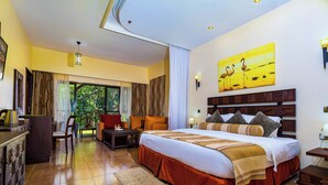 Um quarto amplo com uma cama grande, uma escrivaninha e uma cadeira. Há uma janela com cortinas, um quadro na parede e vista para o verde lá fora.