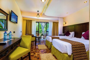 Quarto de hotel com duas camas, uma escrivaninha com cadeira, televisão e janela com cortinas.