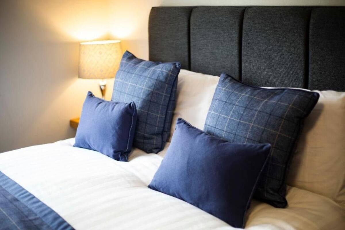 Cama bem arrumada, com cabeceira cinzenta e roupa de cama azul e branca.