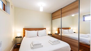 Quarto de hotel com uma cama grande, cabeceira de madeira, mesa de cabeceira com abajur, espelho e janela.