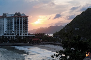 Um hotel à beira-mar com vários andares, uma praia ao primeiro plano e montanhas ao fundo.