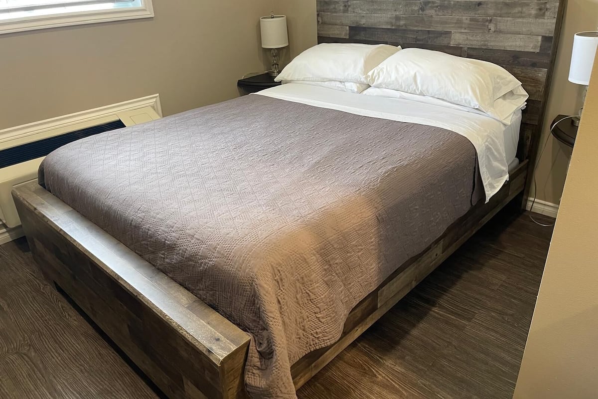 Quarto com estrutura de cama de madeira, colcha cinza e mesa de cabeceira com abajur.