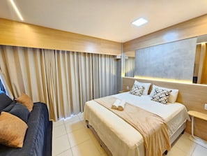 Quarto de hotel moderno com uma cama grande, uma mesinha de cabeceira, um sofá e um espelho amplo.