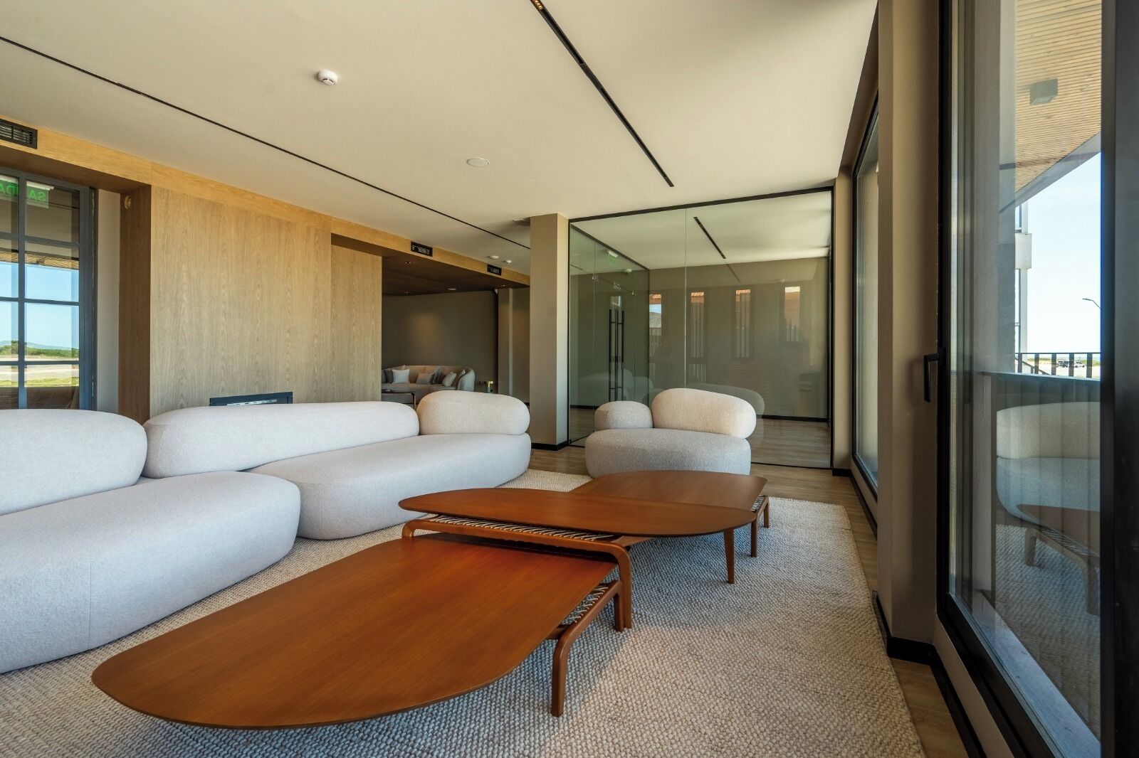 Uma sala de estar moderna com um sofá seccional, duas mesas de centro de madeira e uma parede de vidro que oferece vista para o exterior.