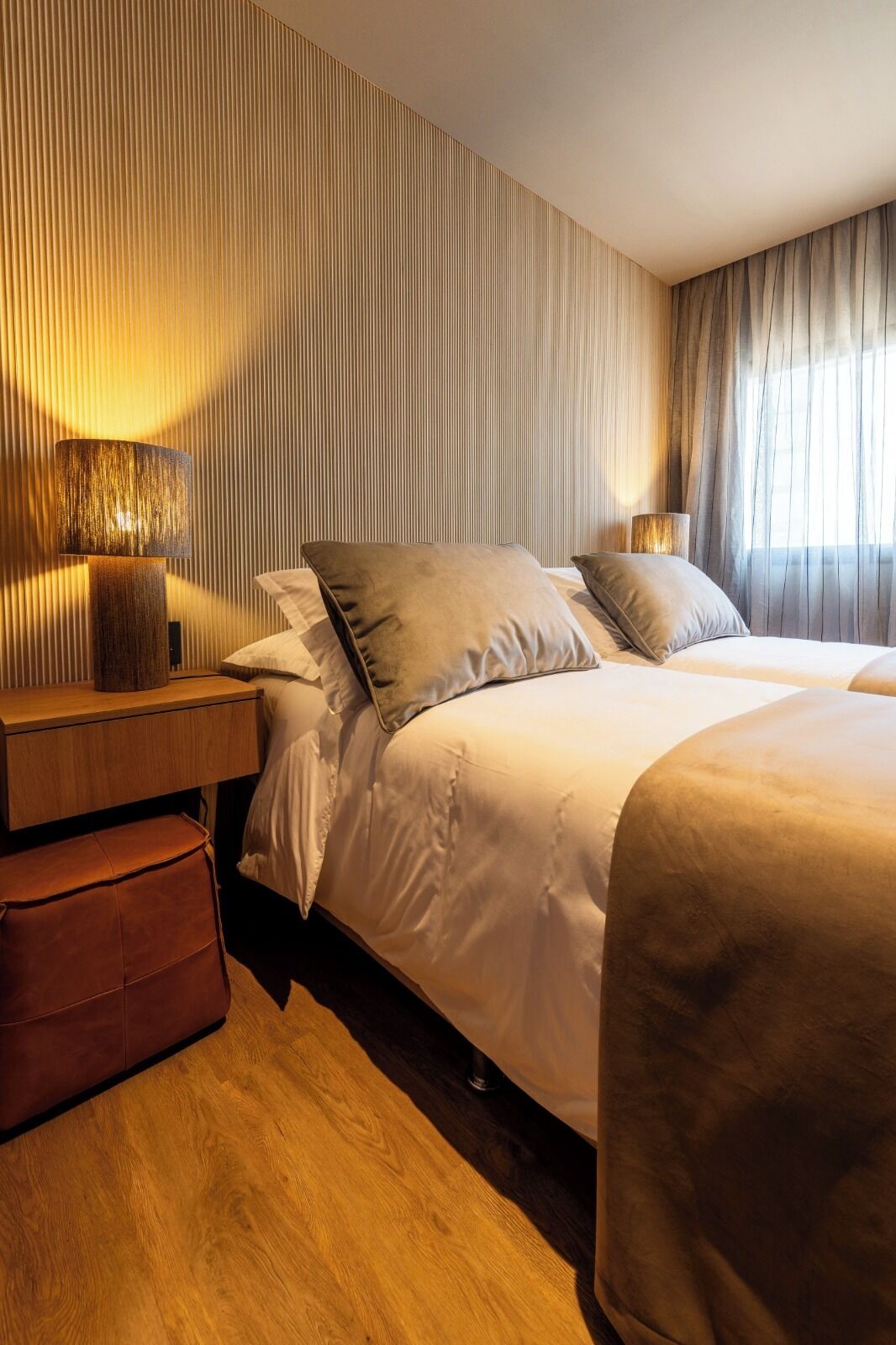 Quarto de hotel moderno com uma cama grande, uma mesa de cabeceira de madeira, um abajur e um banquinho de couro.