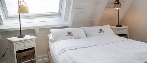 Quarto com claraboia, duas mesas de cabeceira com luminárias, cama com roupa de cama branca e uma pequena mesa de apoio branca com luminária.