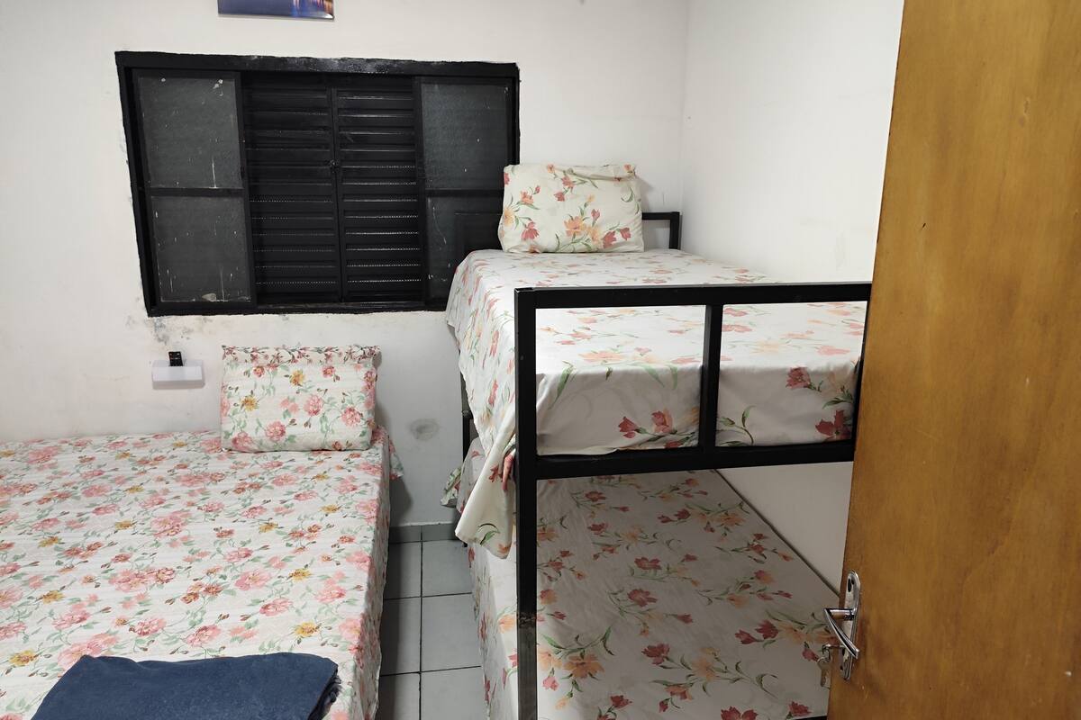 Um pequeno alojamento de um quarto, com duas camas, uma janela com persianas, uma prateleira fixada na parede e uma porta.