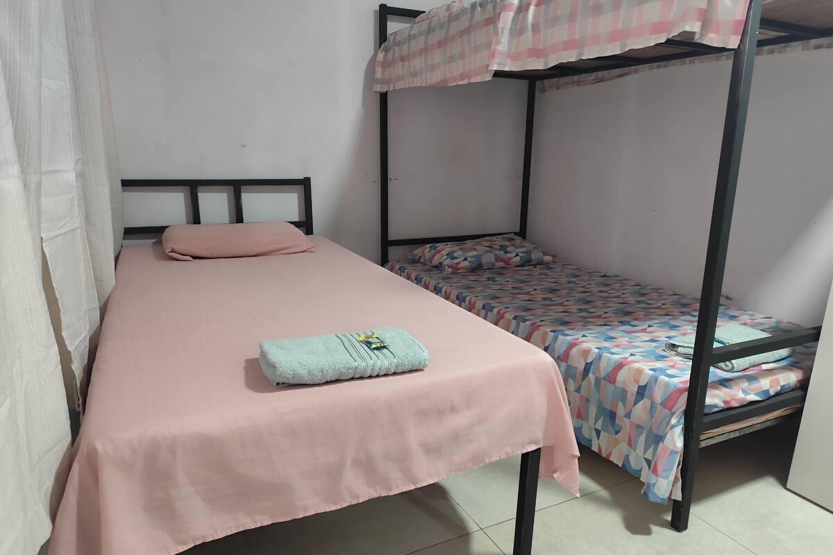 Uma cama de casal com colcha rosa, uma cama menor com colcha estampada e uma cama beliche com dossel xadrez.