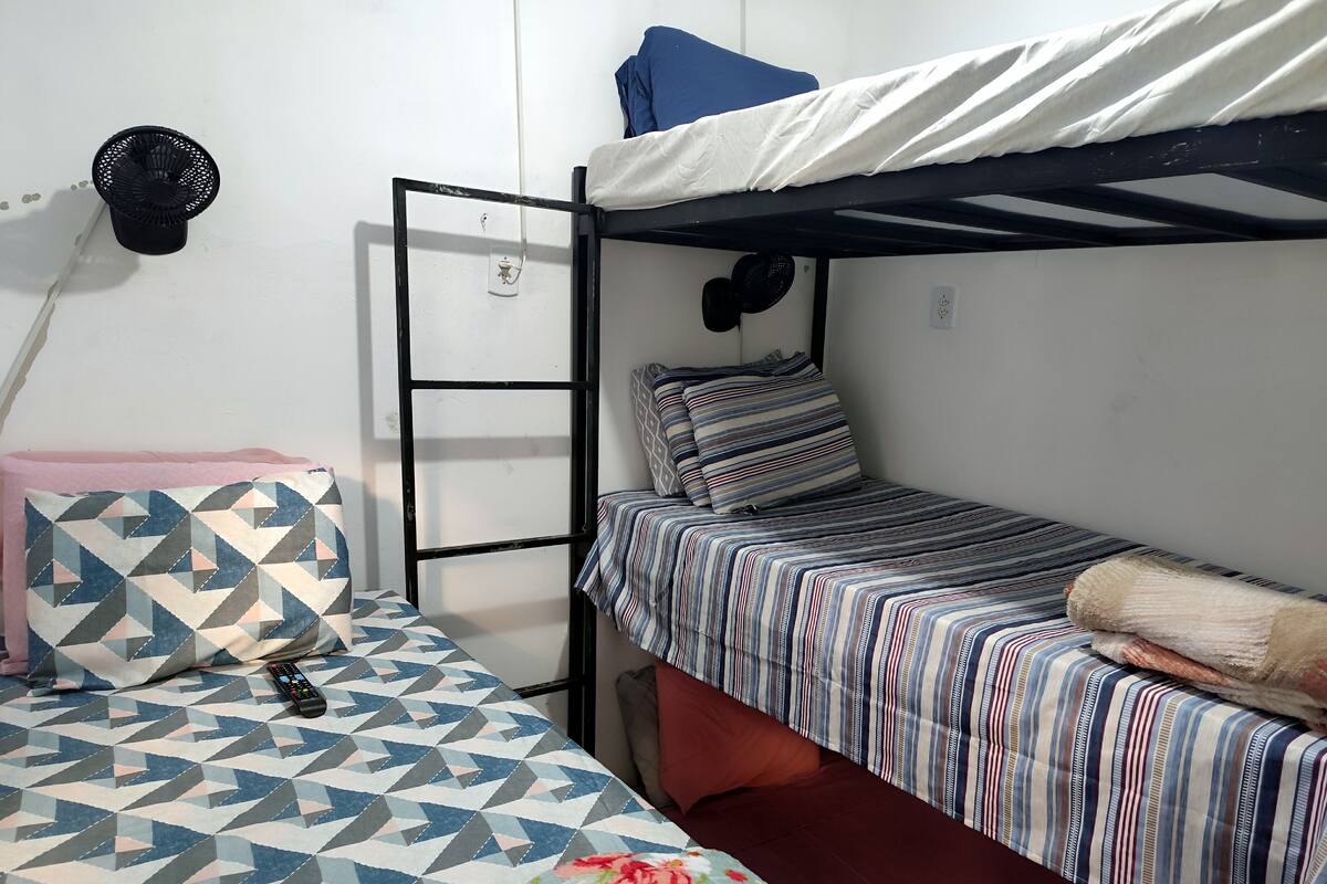 Uma cama beliche com colcha listrada, um travesseiro estampado com padrão geométrico, um controle remoto e um ventilador fixado na parede.