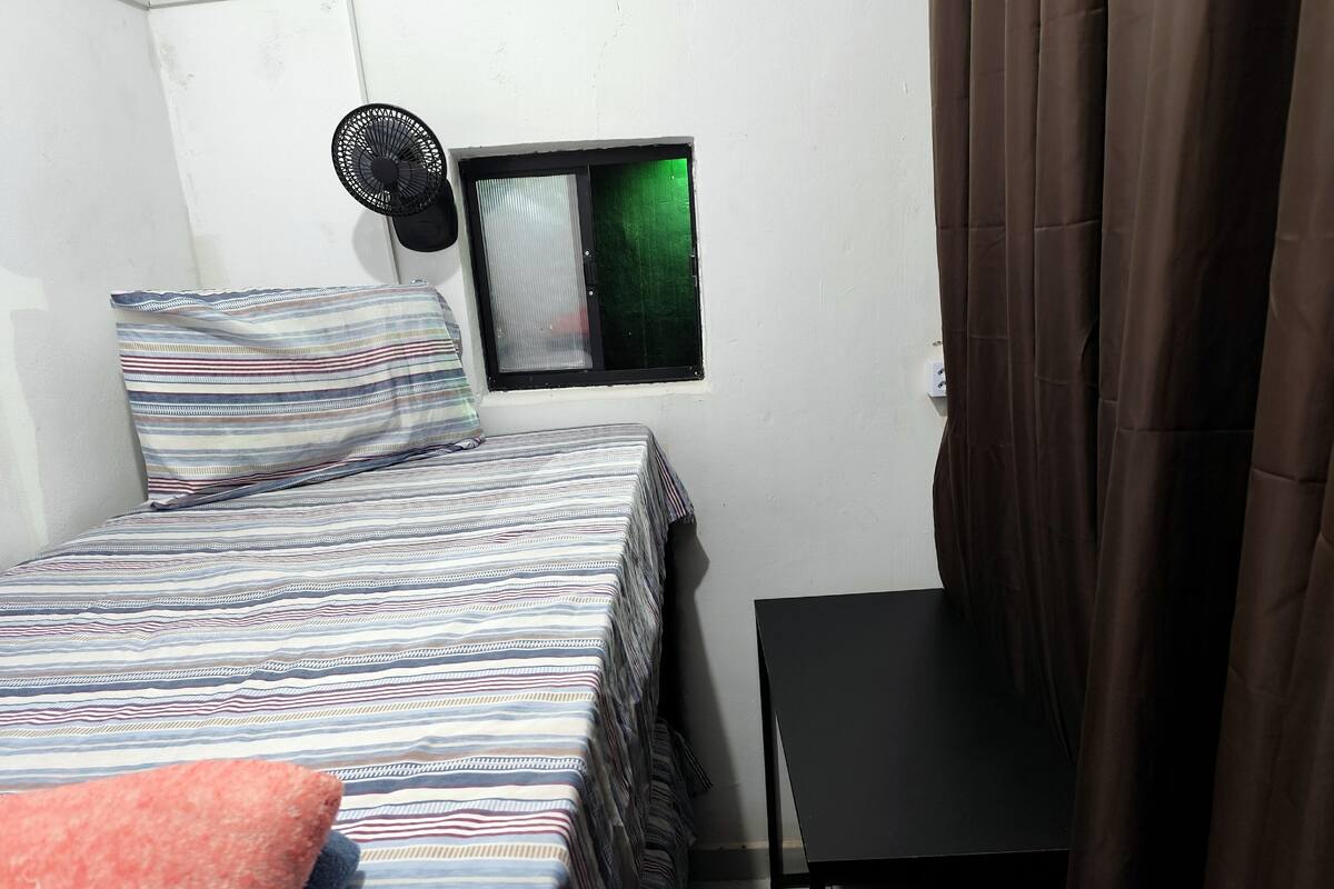 Quarto com cama, uma mesinha, um ventilador, janela com vista para o verde e cortina.