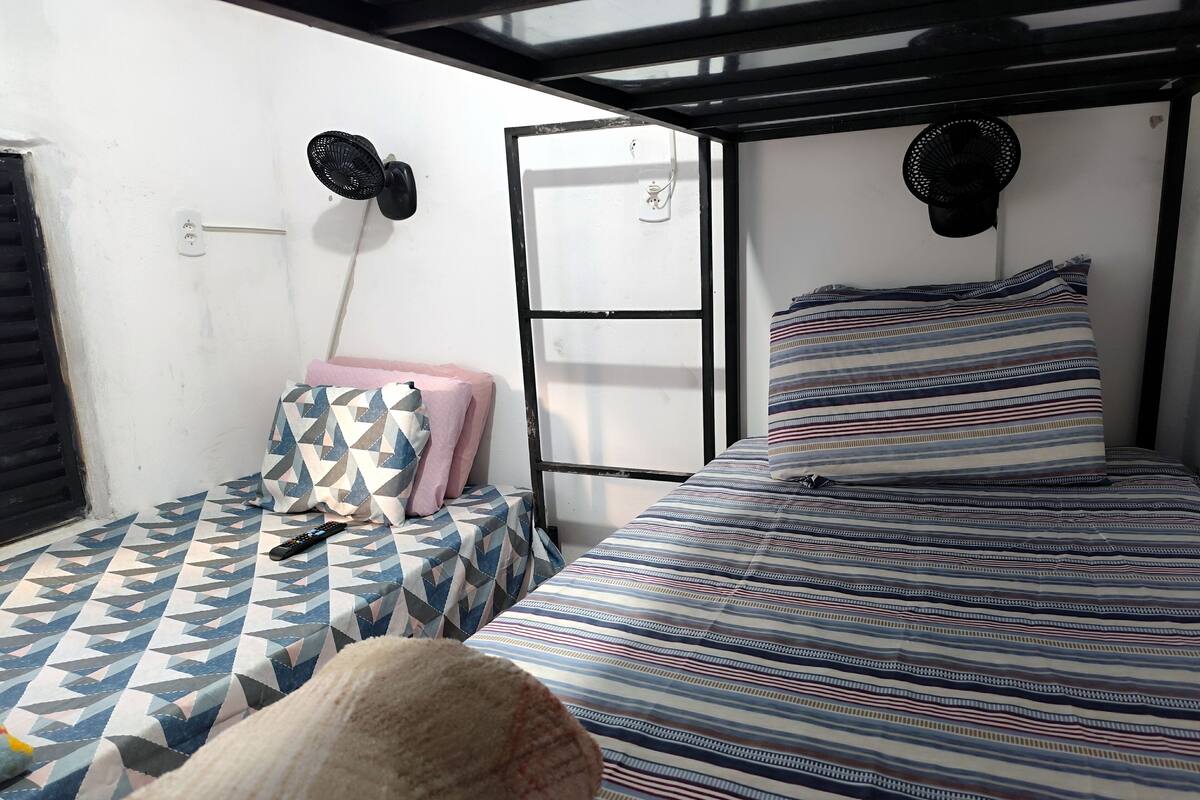 Quarto com duas camas, uma com colcha de padrão geométrico e outra com colcha listrada. Há dois ventiladores fixados na parede, um sobre cada cama.